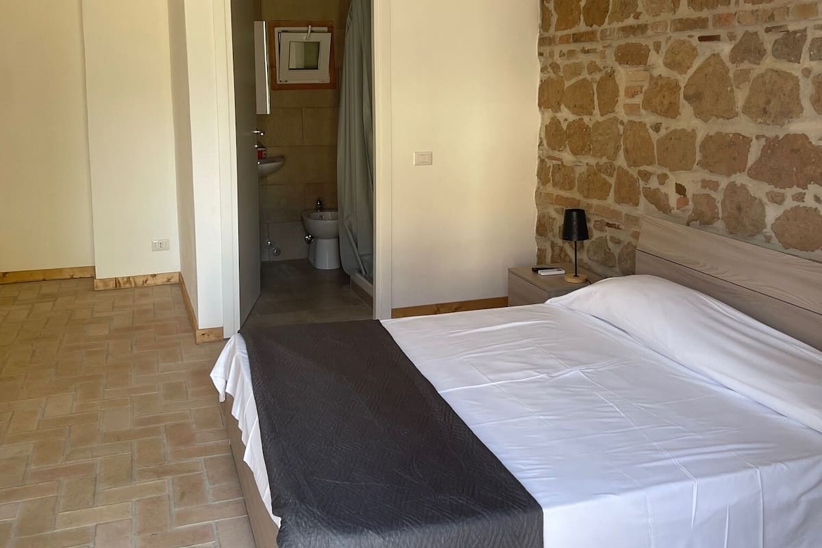 Quarto com parede de pedra, cama com roupa de cama branca, mesa de cabeceira com abajur e banheiro visível por uma porta aberta.
