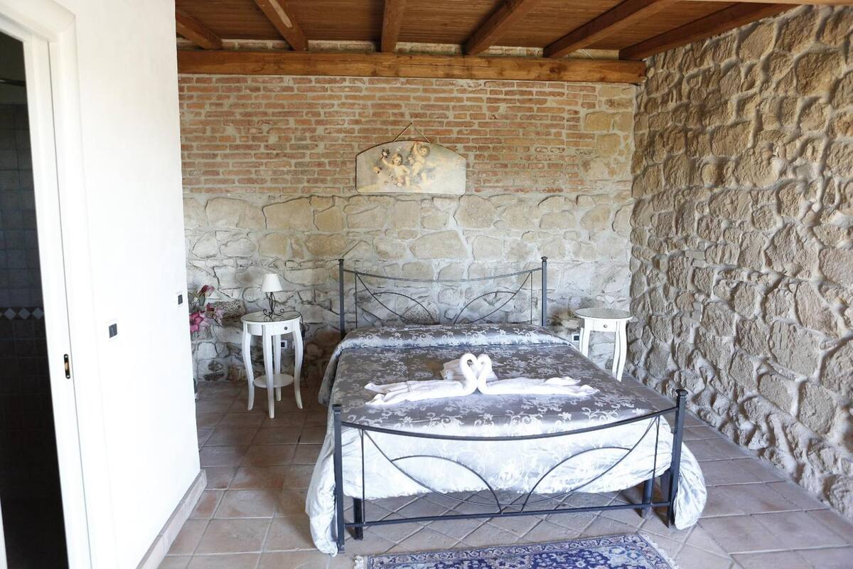 Quarto com parede de pedra, estrutura de cama de metal e mesinha de cabeceira com abajur.