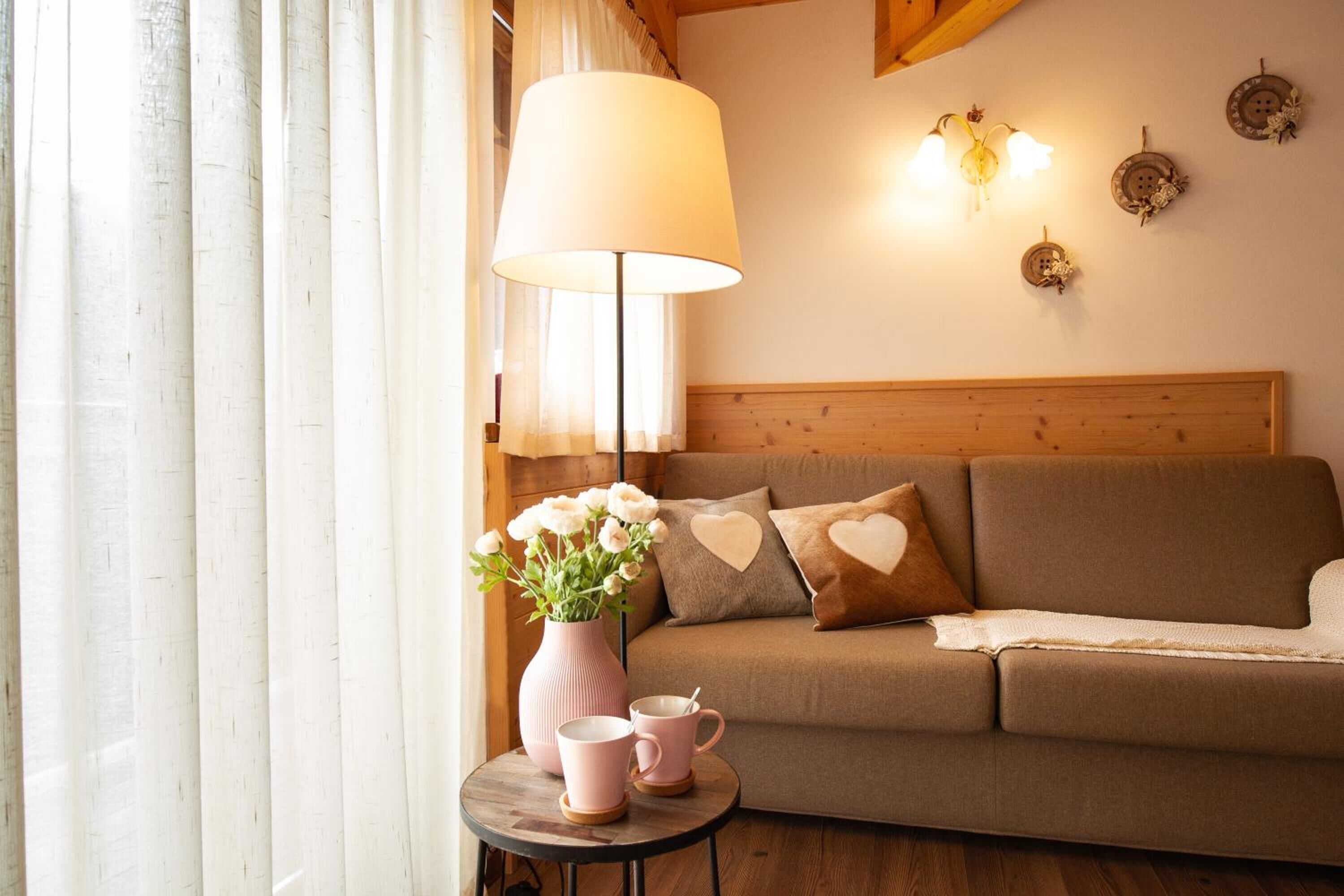 Quarto aconchegante com cabeceira de madeira, sofá, mesa com um vaso de flores e duas canecas cor-de-rosa, e um abajur.