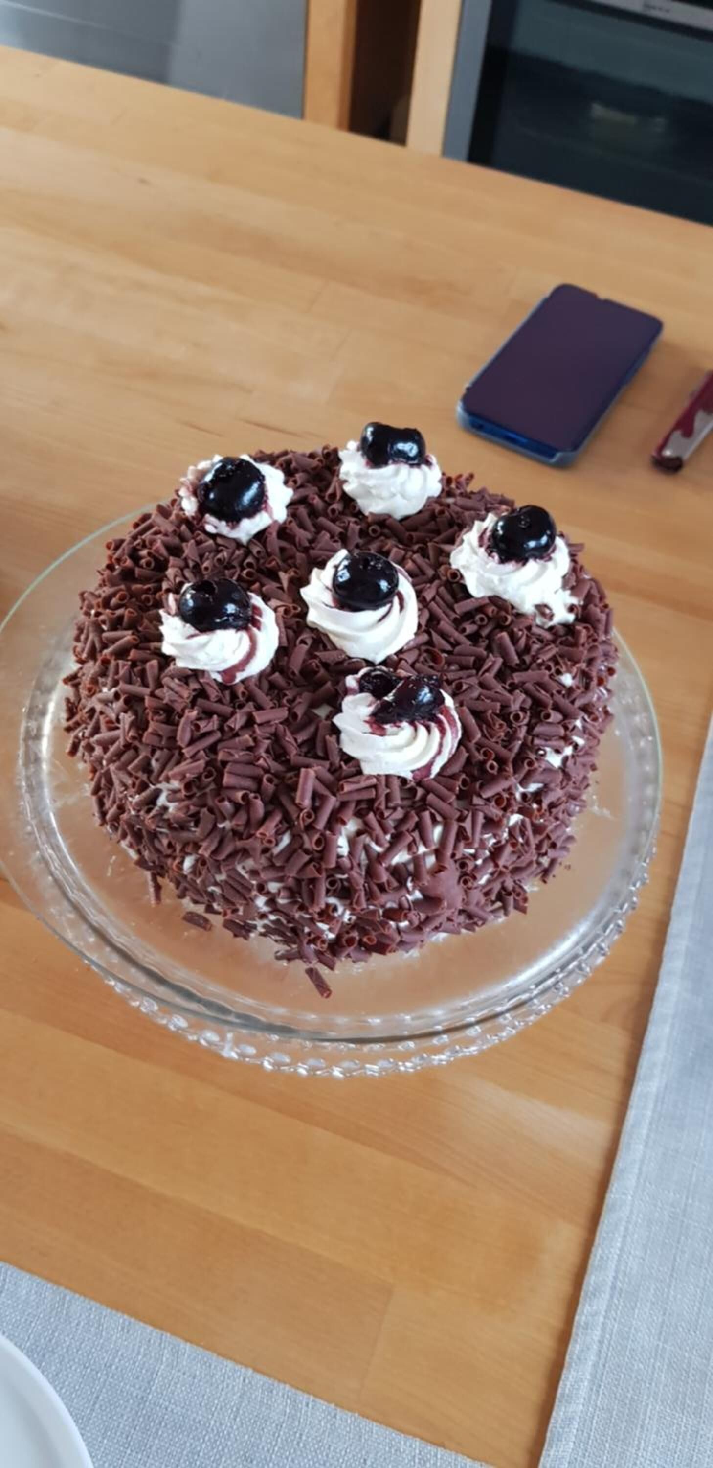 Bolo de chocolate redondo com chantilly e cerejas sobre um prato de vidro em uma mesa de madeira.