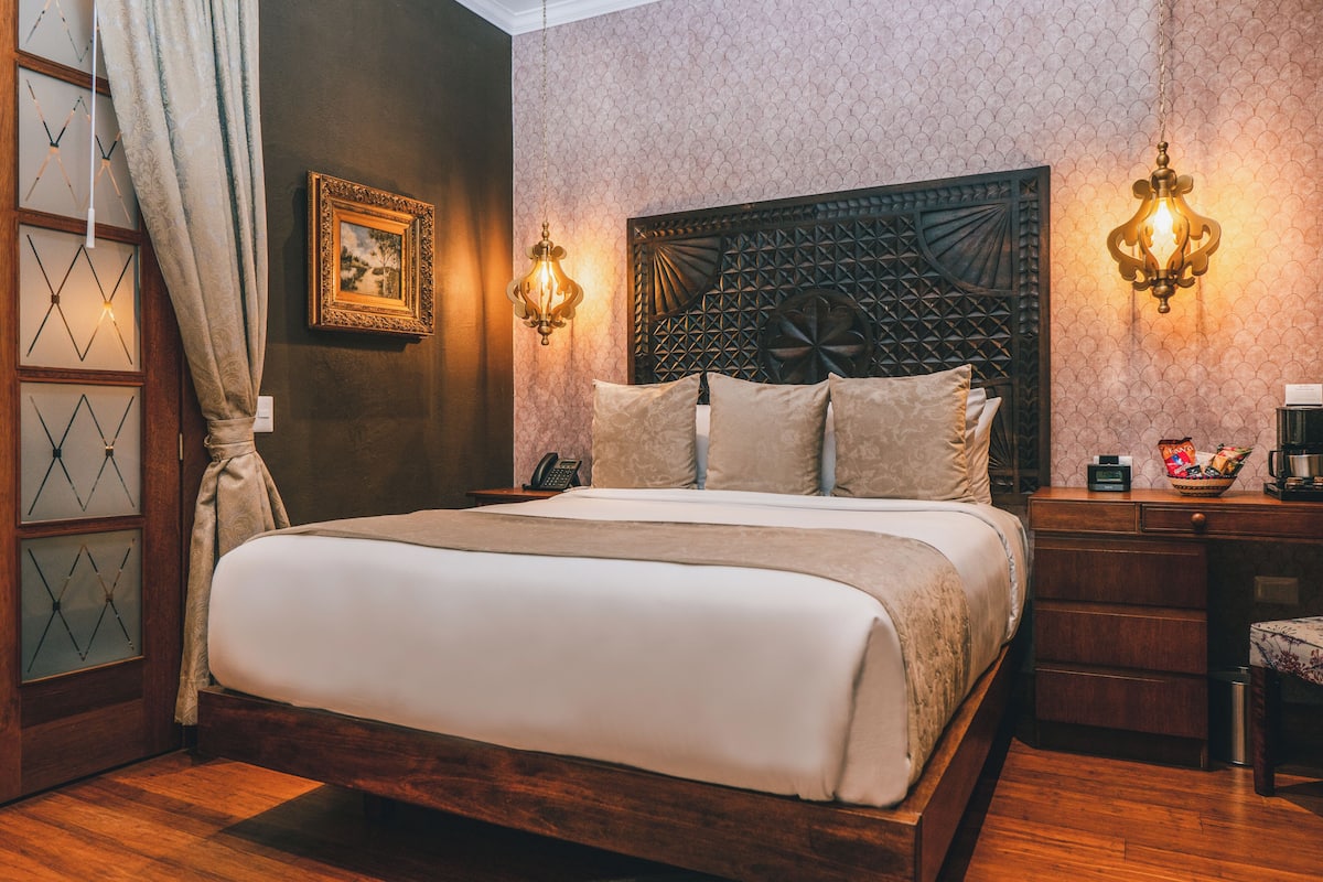 Uma cama bem arrumada com cabeceira, abajures laterais, telefone ao lado da cama e mesa de cabeceira com cafeteira e cesta.