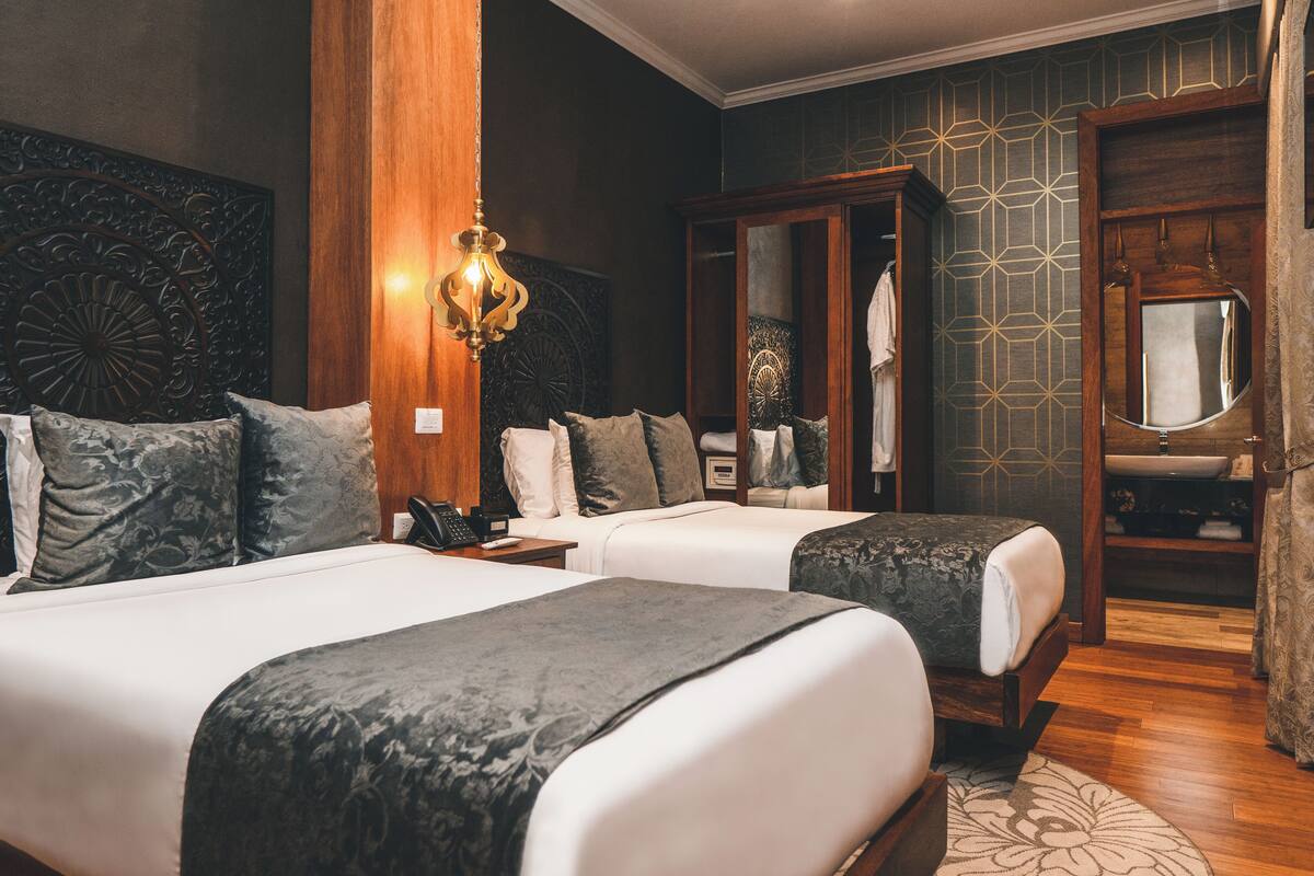 Quarto de hotel com duas camas, um guarda-roupa de madeira, um espelho e um papel de parede estampado.