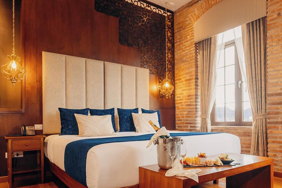 Quarto de hotel com uma cama grande, uma mesinha de cabeceira, uma mesa de centro de madeira e uma janela com cortinas.