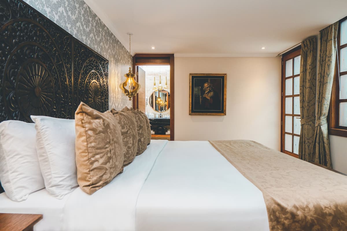 Cama bem arrumada com colcha estampada e almofadas, parede com cabeceira de metal decorativa, quadro emoldurado e janela com cortinas.