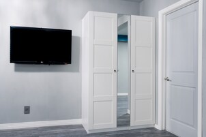 Quarto com televisão de tela plana na parede, guarda-roupa branco com porta espelhada e porta branca fechada.