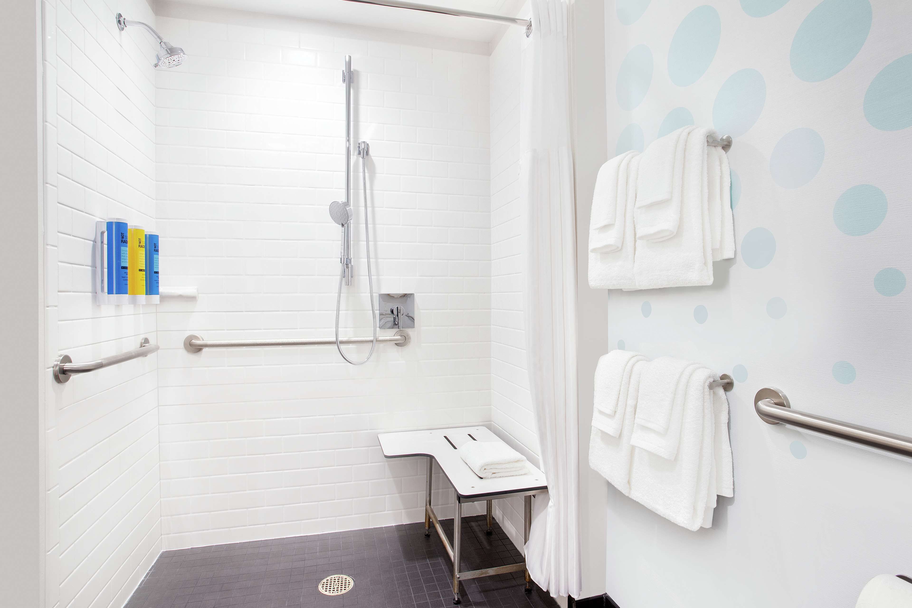 Um banheiro moderno com azulejos brancos estilo metrô, chuveiro com chuveiro manual, barra de apoio e porta-toalhas com vários toalhas.