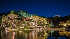 Uma paisagem noturna tranquila com um lago refletor, árvores iluminadas e um edifício de estilo japonês tradicional ao fundo.