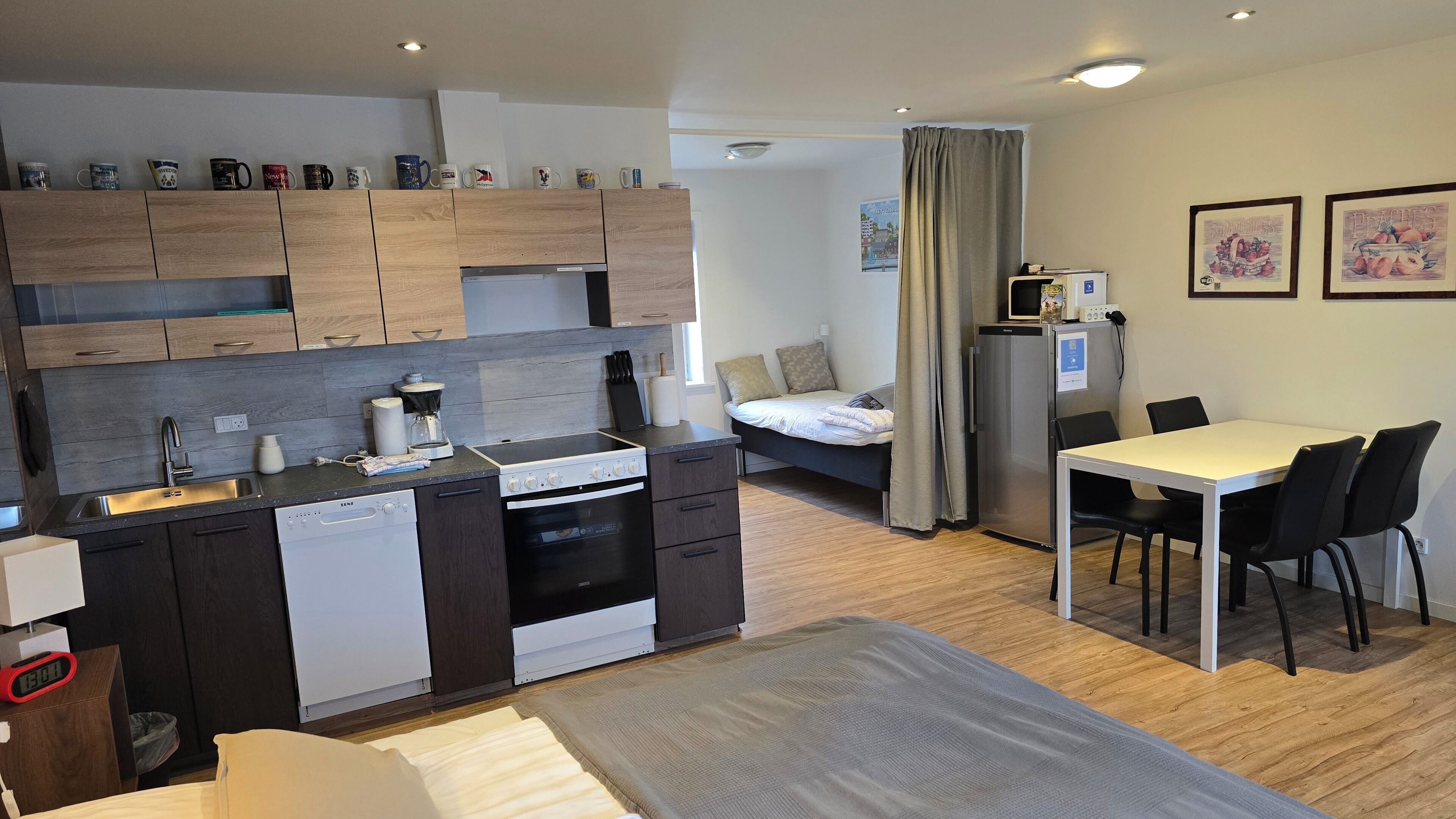 Cozinha compacta com lava-louças, fogão e máquina de café. Há uma área de refeições com mesa e cadeiras, e uma cama com cortina.