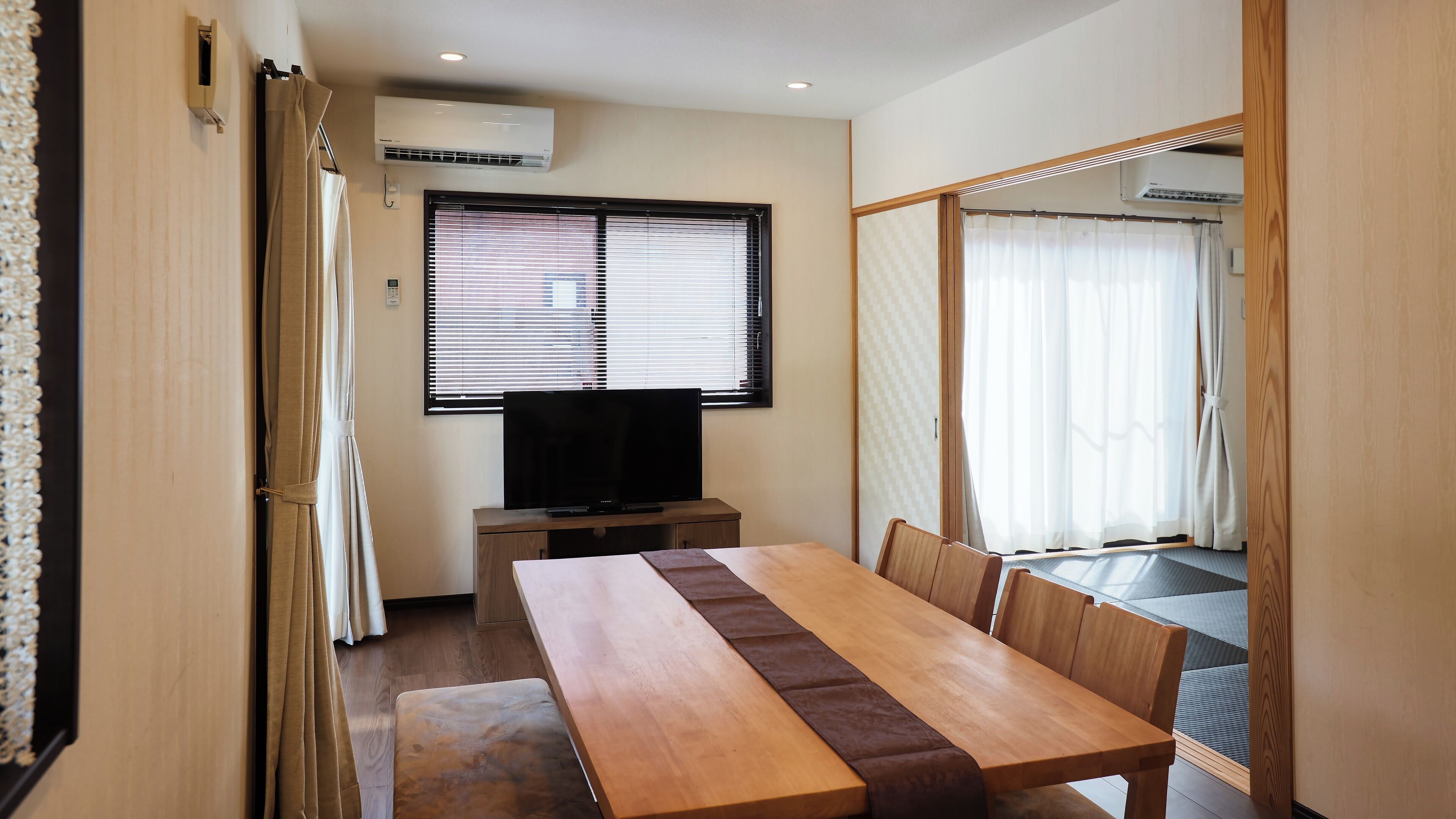 Quarto com uma mesa de jantar de madeira, uma televisão em móvel e uma janela com persianas.