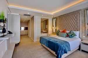 Quarto moderno com cama grande, cabeceira estofada, mesa de cabeceira e mesa de apoio. Há uma escrivaninha com cadeira e um espelho ao fundo.
