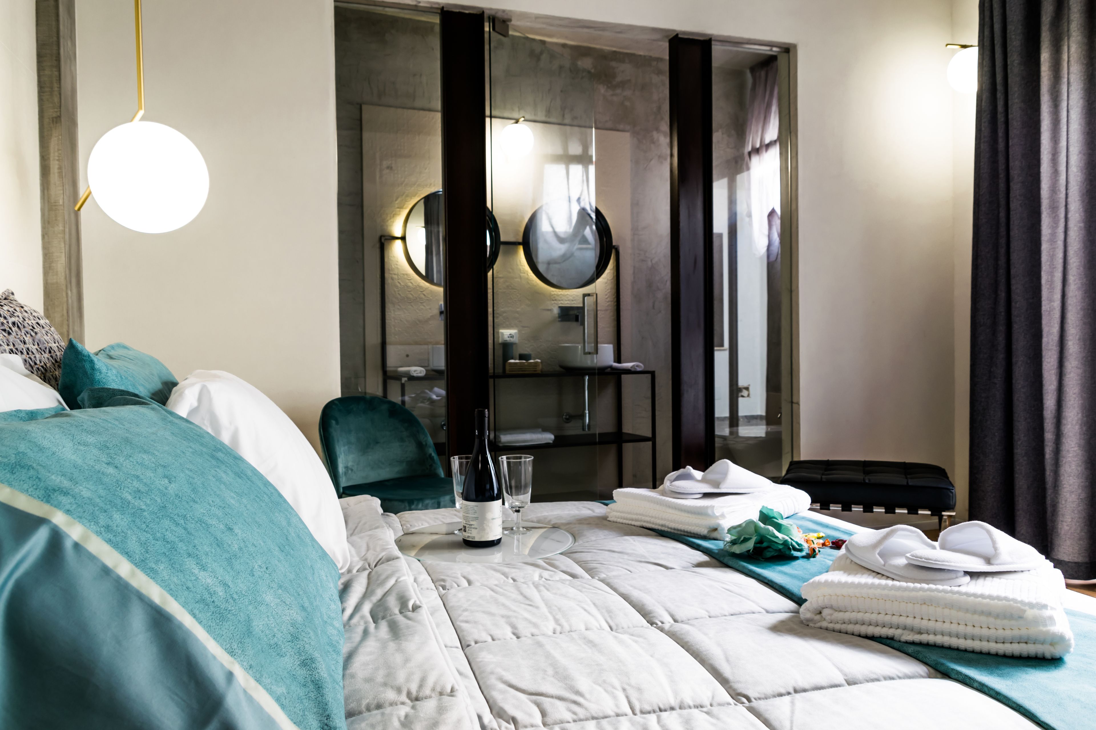 Quarto de hotel com cama, mesa de cabeceira com garrafa e copos, espelho, um banco e janela com cortinas.