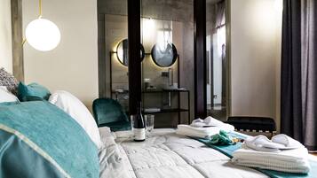 Quarto de hotel com cama, mesa de cabeceira com garrafa e copos, espelho, um banco e janela com cortinas.