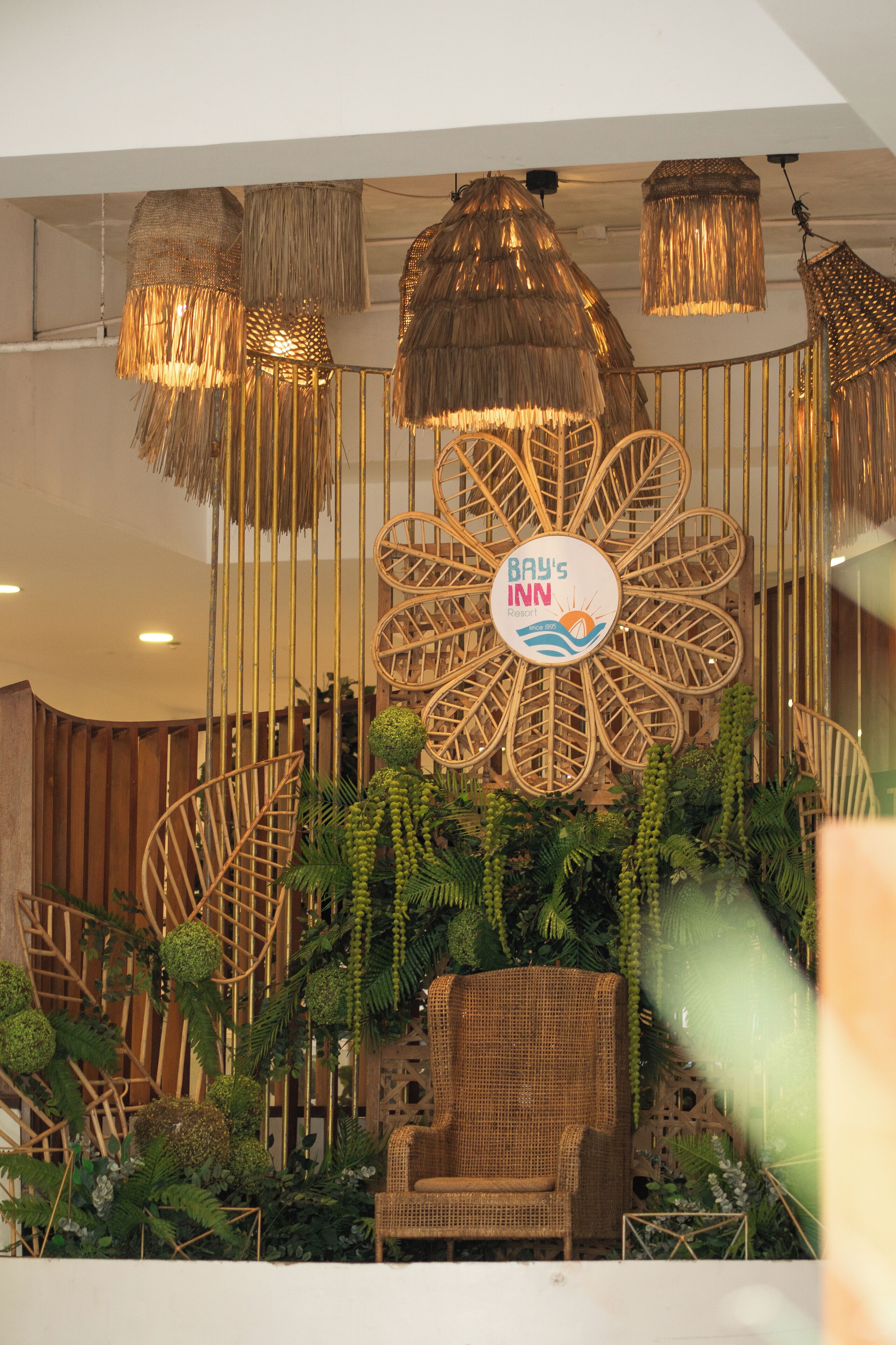 Área de lobby com uma estrutura decorativa central, uma cadeira de vime e luminárias pendentes.