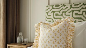 Cama bem arrumada com almofadas estampadas, mesa de cabeceira com um vaso e um copo de vidro, e uma luminária na parede.