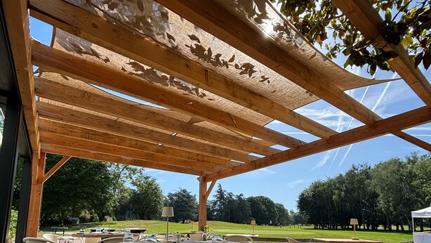 Um pátio externo com mesas e cadeiras de madeira, uma pérgola acima e vista para um parque.