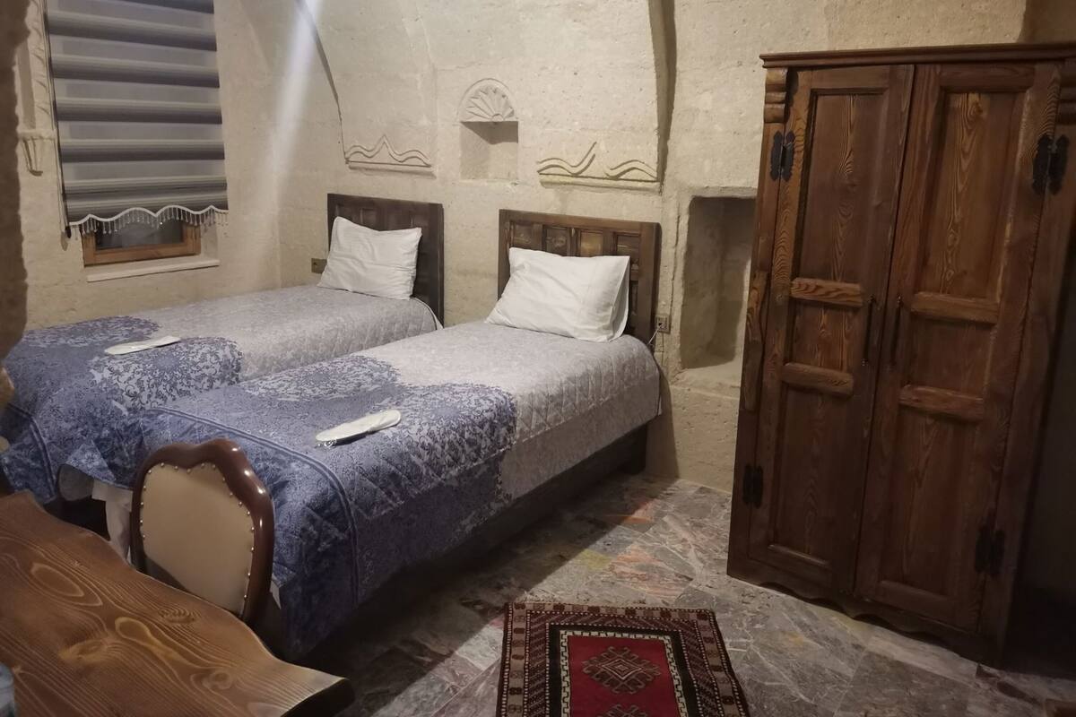 Quarto com duas camas, um guarda-roupa de madeira, uma mesinha e uma janela com cortina.