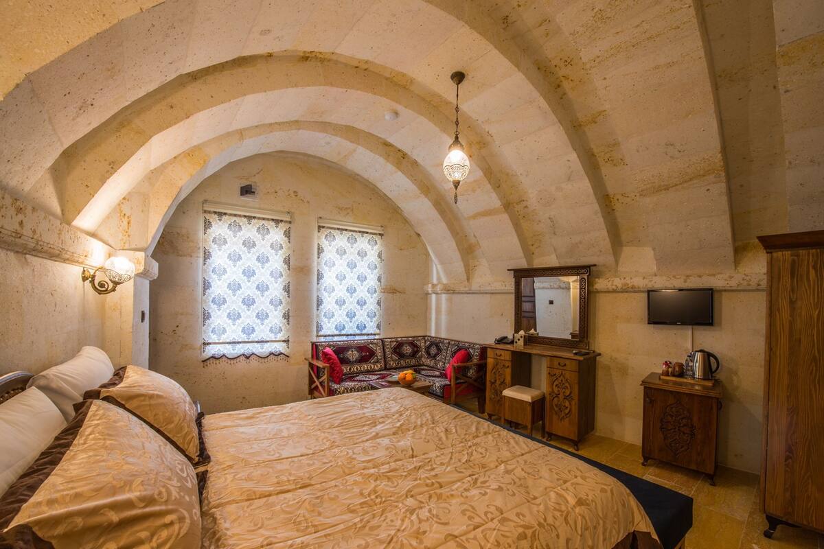 Quarto com cama grande, cómoda de madeira, televisão e janela com cortinas estampadas.