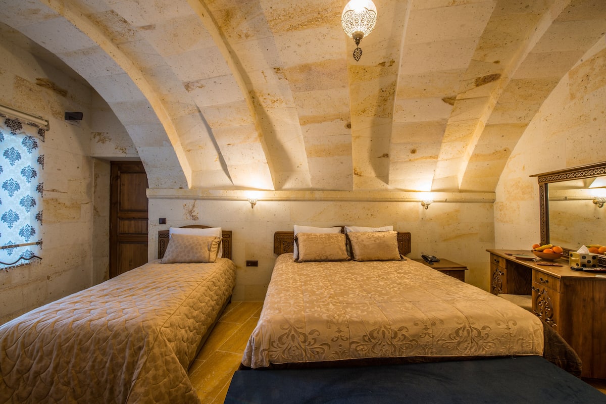 Quarto com duas camas, um aparador de madeira, um espelho e uma janela com cortinas estampadas.