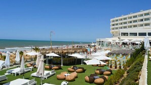 Um hotel à beira-mar com áreas de estar ao ar livre, incluindo espreguiçadeiras e guarda-sóis, e uma área de piscina com cobertura de palha.