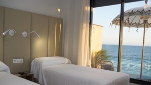 Quarto de hotel com duas camas, uma janela grande com vista para o mar e um ventilador de teto.