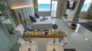 Quarto de hotel moderno com cama, mesas de cabeceira, toucador com espelho, pequena mesa com artigos de higiene e vista para o mar.