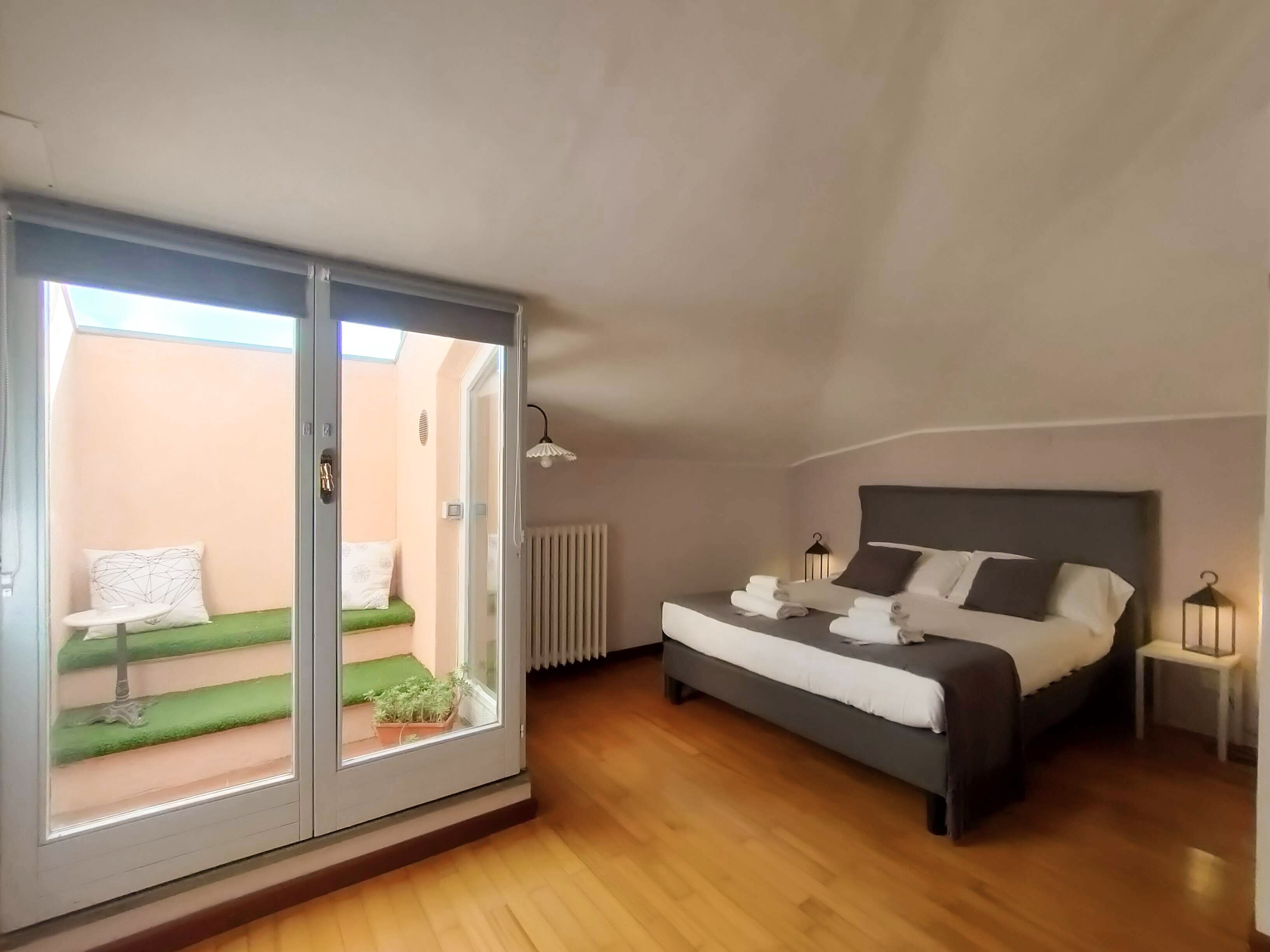 Quarto com cama, mesa de cabeceira, luminária e porta de vidro que desliza para uma varanda com cadeira e pequena mesa.