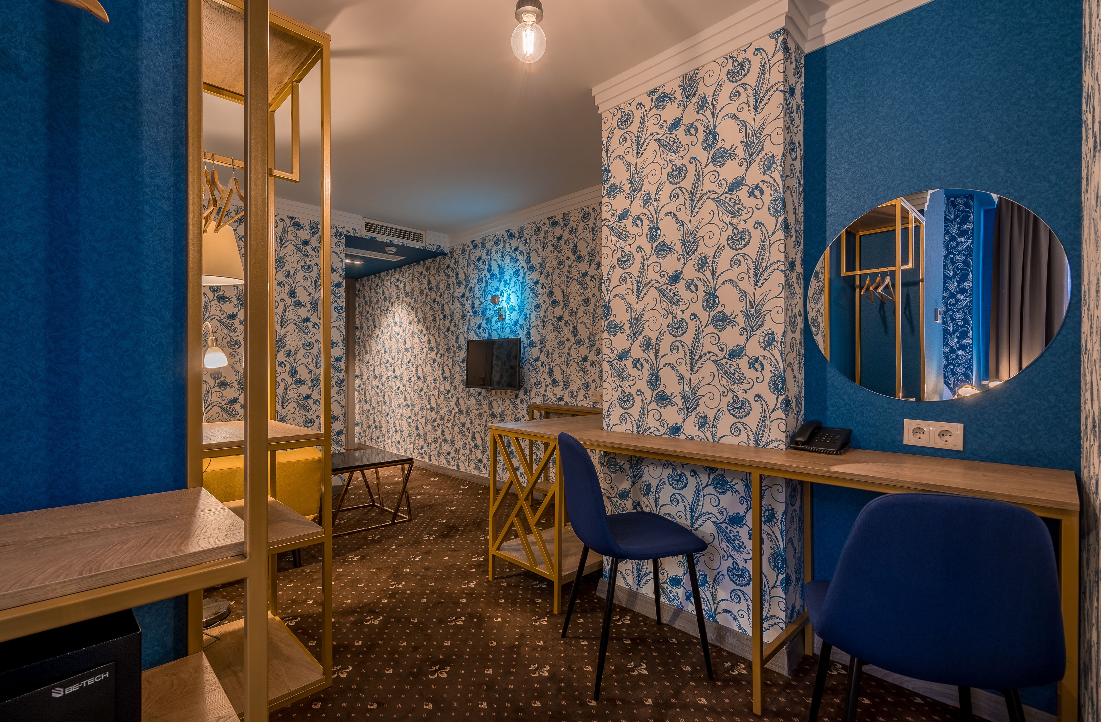 Quarto com papel de parede estampado em azul e branco, um espelho redondo, uma escrivaninha com uma cadeira e um lustre.