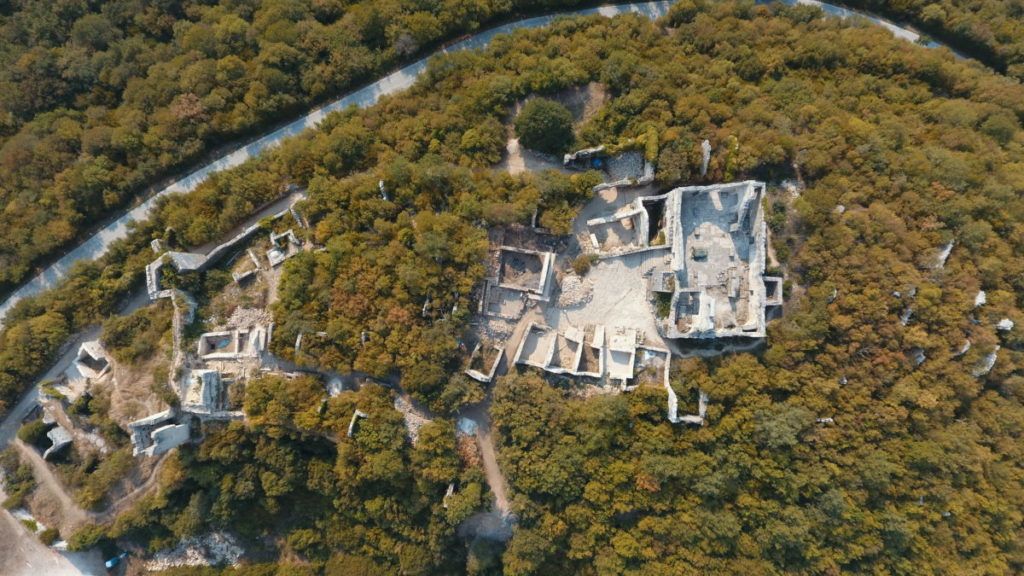 Vista aérea de ruínas antigas com várias estruturas, cercadas por uma floresta densa.