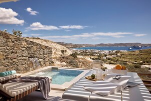 Piscina com muro de pedra, cadeiras de descanso, mesa com livro e frutas, e vista para o mar e colinas.