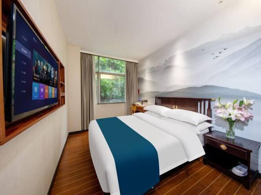 Quarto de hotel com cama, televisão na parede, mesa de cabeceira e janela com cortinas.