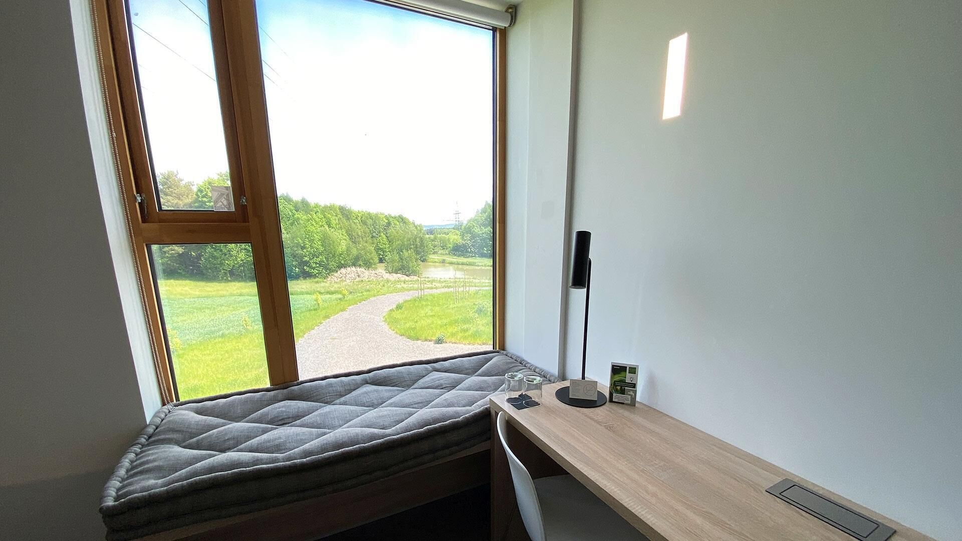 Quarto com cama, escrivaninha e janela com vista para uma paisagem verde.