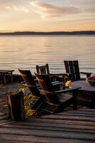Um deck de madeira com cadeiras Adirondack e uma mesa, com vista para um corpo d'água calmo ao pôr do sol.