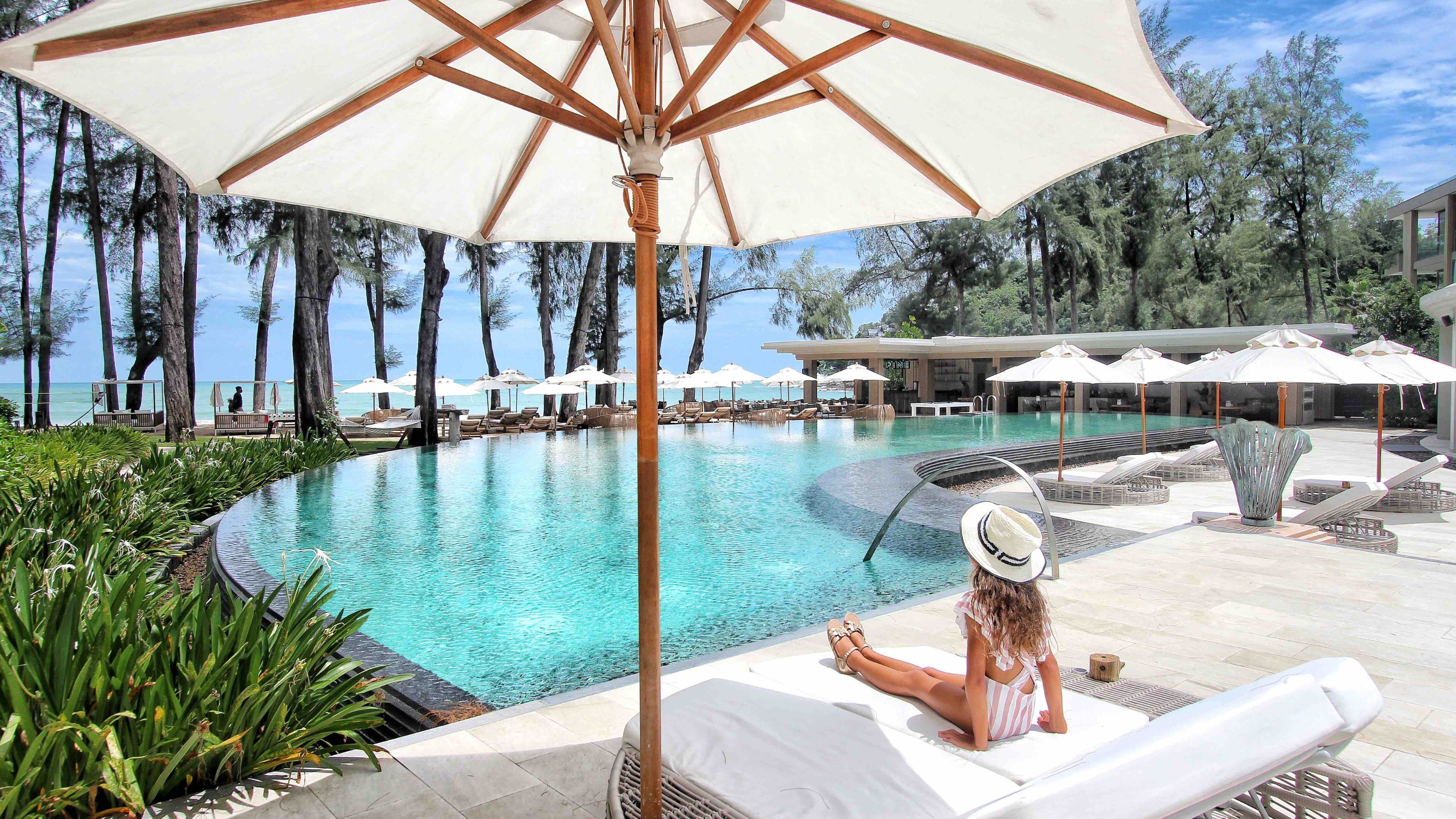 Área da piscina com espreguiçadeiras e guarda-sóis, cercada por árvores e vista para o mar.