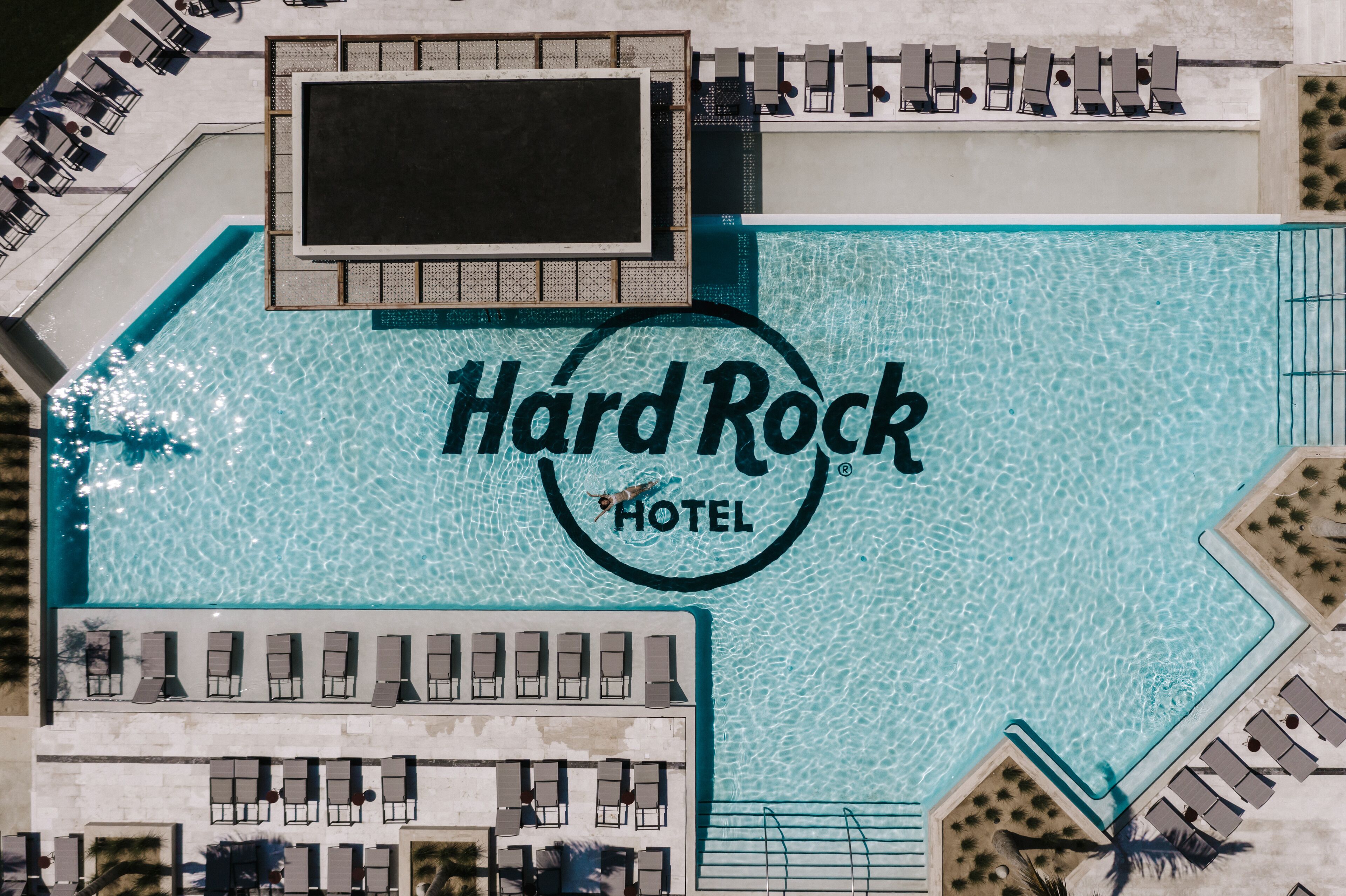 Vista aérea de um hotel com uma grande piscina, áreas de lazer e um logotipo central.