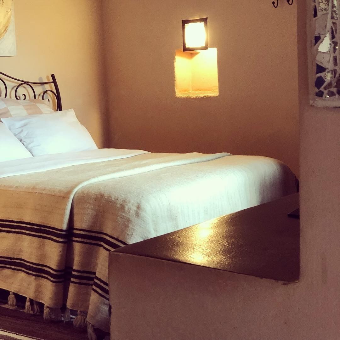 Uma cama com edredom branco e manta listrada, luminária fixada na parede e janela com moldura decorativa.