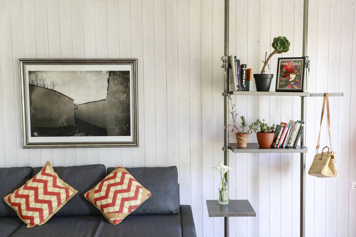 Sala de estar com um sofá cinzento, duas almofadas estampadas, um quadro abstrato emoldurado, uma mesinha de apoio com um vaso de flores brancas, uma estante com livros e plantas, e uma bolsa pendurada.
