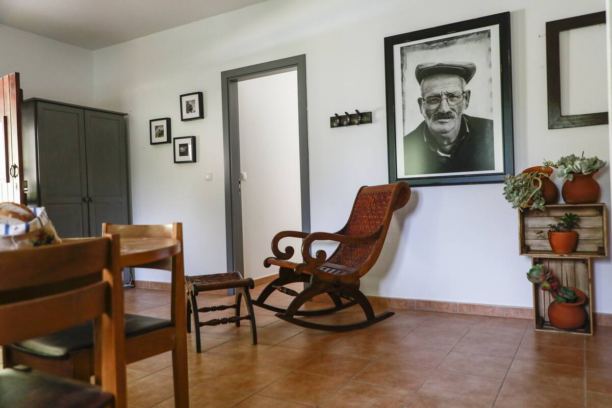 Quarto com uma cadeira de balanço, uma cadeira de madeira, uma porta e uma parede com quadros e um retrato grande.