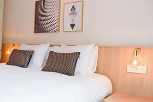 Cama bem arrumada com almofadas brancas e estampadas, cabeceira de madeira e duas luminárias fixadas na parede.