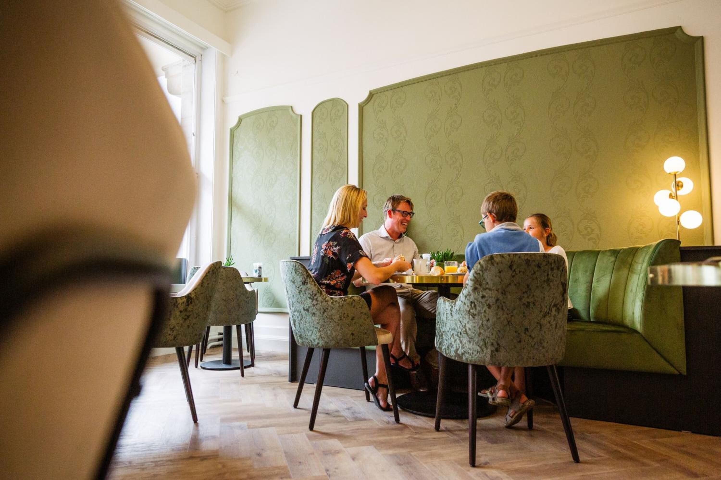 Uma sala de jantar com um sofá estofado verde, cadeiras estampadas e uma mesa com pessoas sentadas.