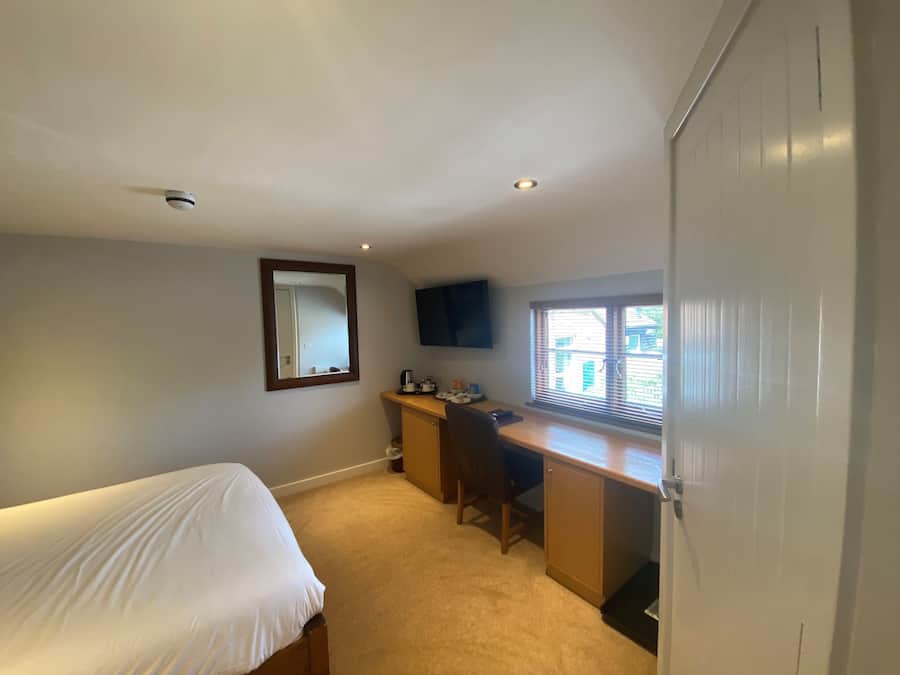 Quarto com cama, escrivaninha, cadeira, televisão e janela.