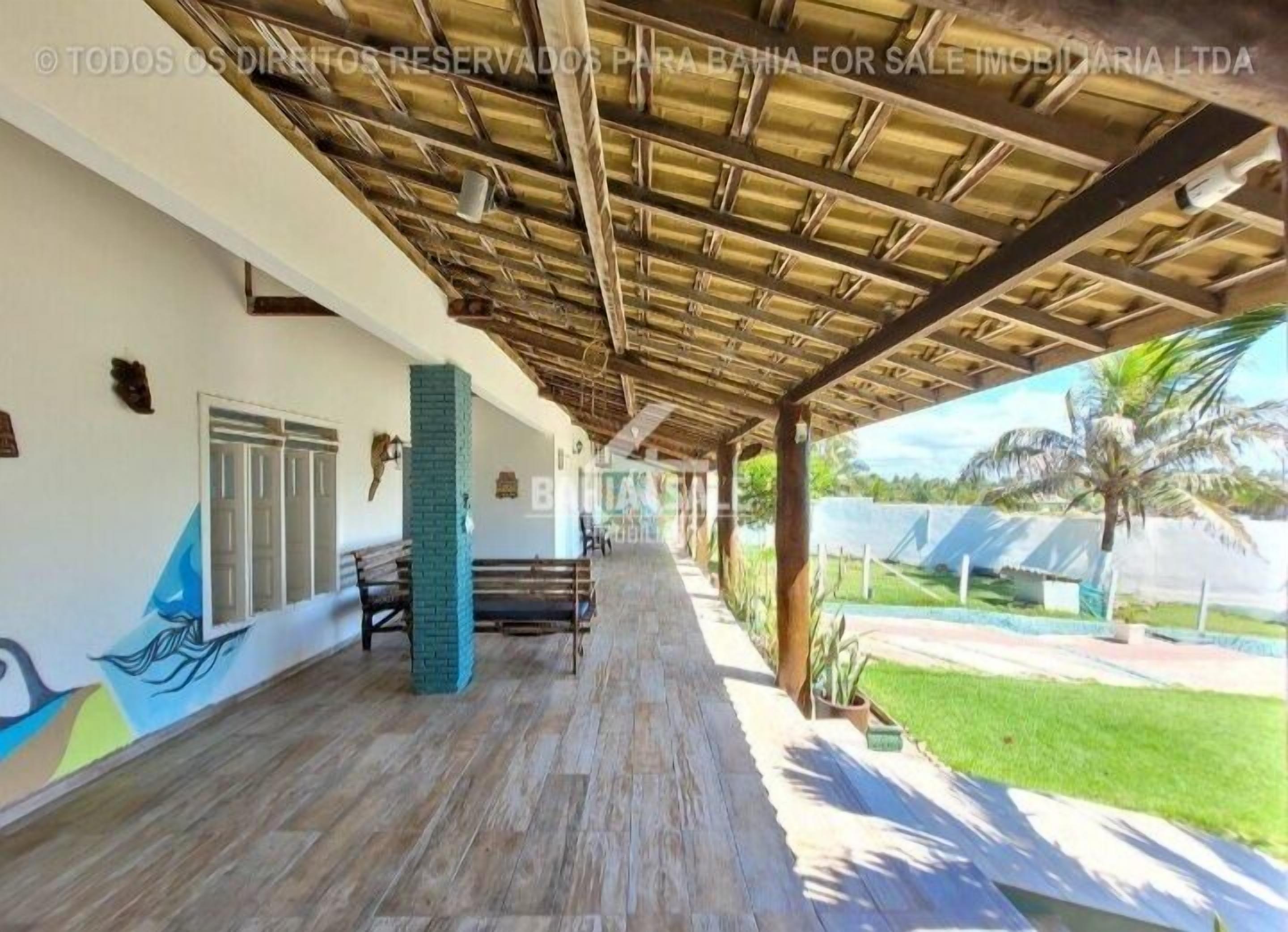 Espaço ao ar livre coberto com piso de madeira, bancos e vista para uma área gramada com palmeiras.