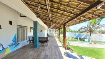 Espaço ao ar livre coberto com piso de madeira, bancos e vista para uma área gramada com palmeiras.