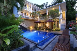 Piscina com deck de madeira, cercada por vegetação exuberante e um edifício com varandas múltiplas.
