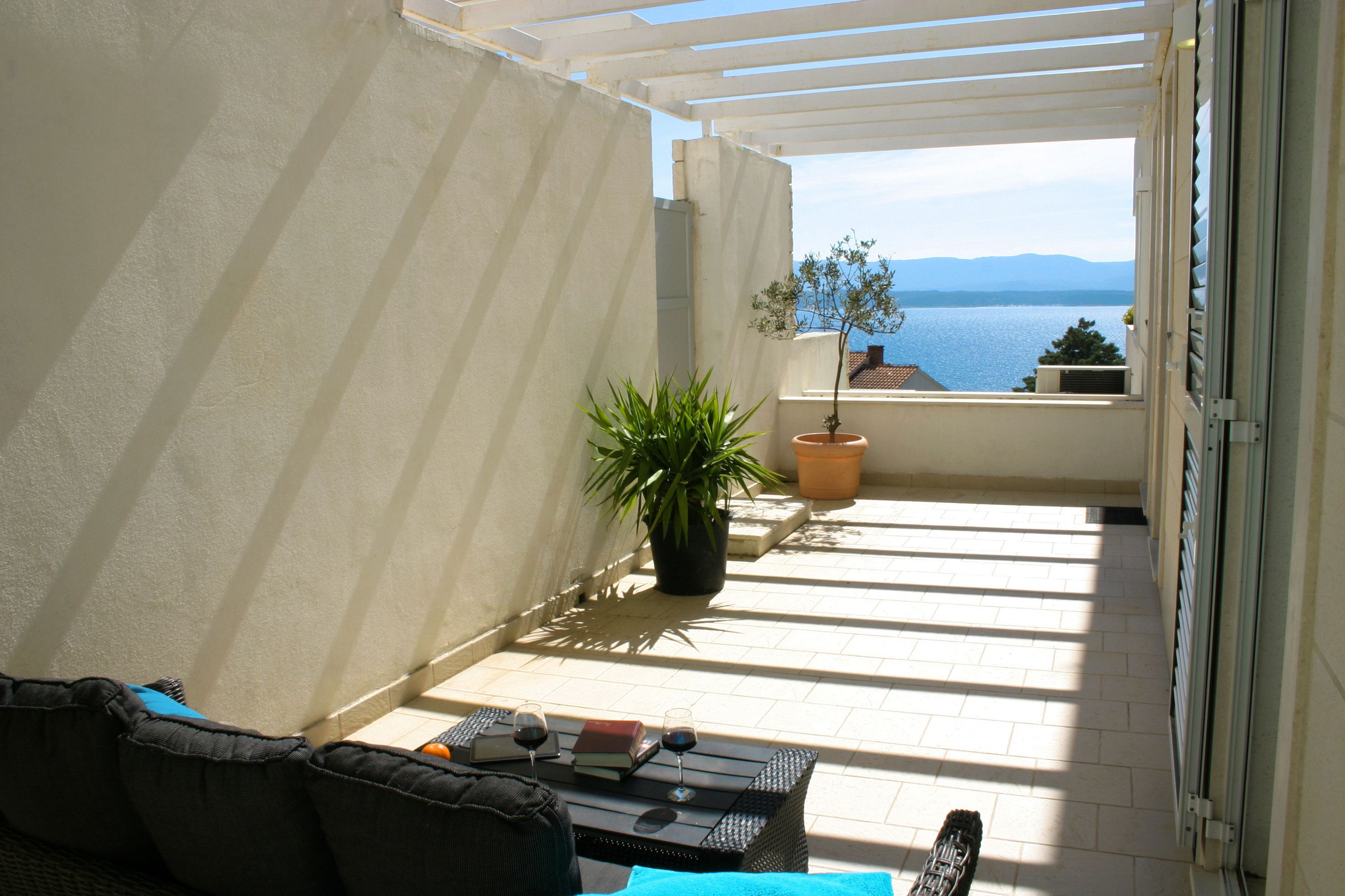 Varanda ensolarada com sofá, mesa com livros e copos, plantas em vasos e vista para o mar.