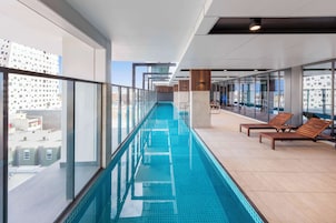 Piscina interior moderna com paredes de vidro transparente, espreguiçadeiras de madeira e vista para a paisagem urbana.