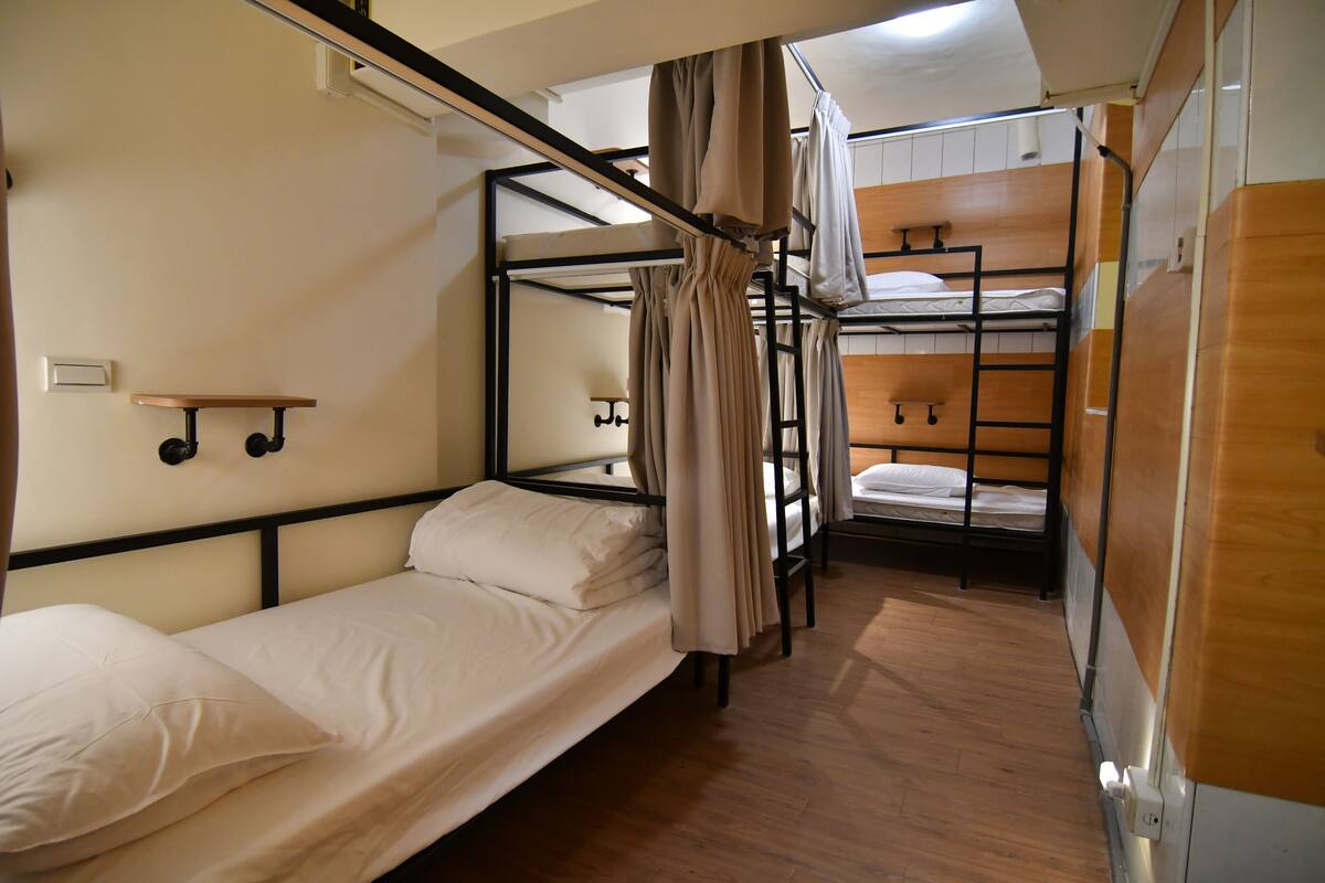 Quarto com beliche, piso de madeira, uma mesinha e prateleira com dois ganchos.