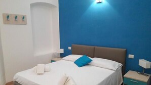 Uma cama bem arrumada com lençóis brancos e um travesseiro azul, uma mesa de cabeceira com abajur, e um luminário fixado na parede.