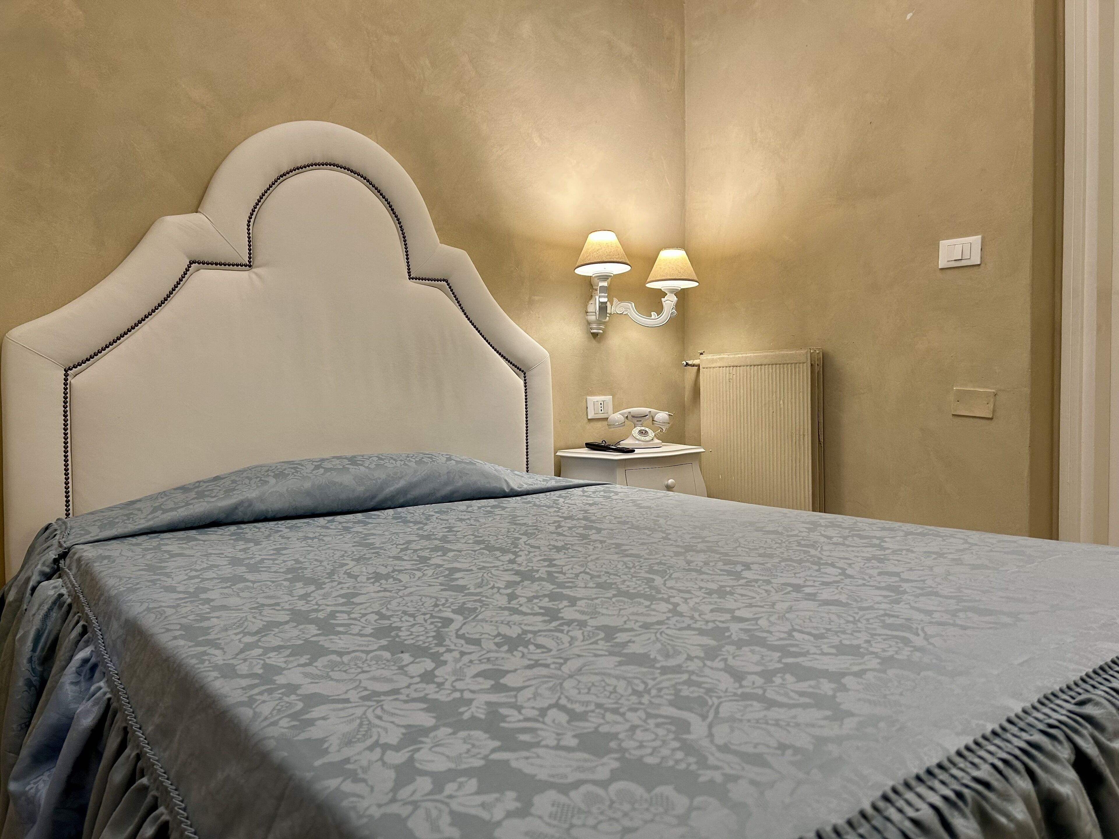 Quarto com cabeceira estofada, mesa de cabeceira com abajur e radiador.