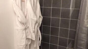 Um banheiro com duas roupões brancos pendurados em ganchos, uma cortina de chuveiro e um par de chinelos no chão.