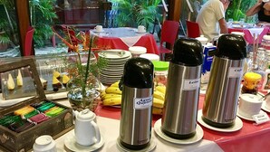 Mesa de buffet de café da manhã com diversos alimentos, incluindo bananas, uma variedade de frutas e garrafões térmicos com os dizeres 'Leite' e 'Água Quente'.